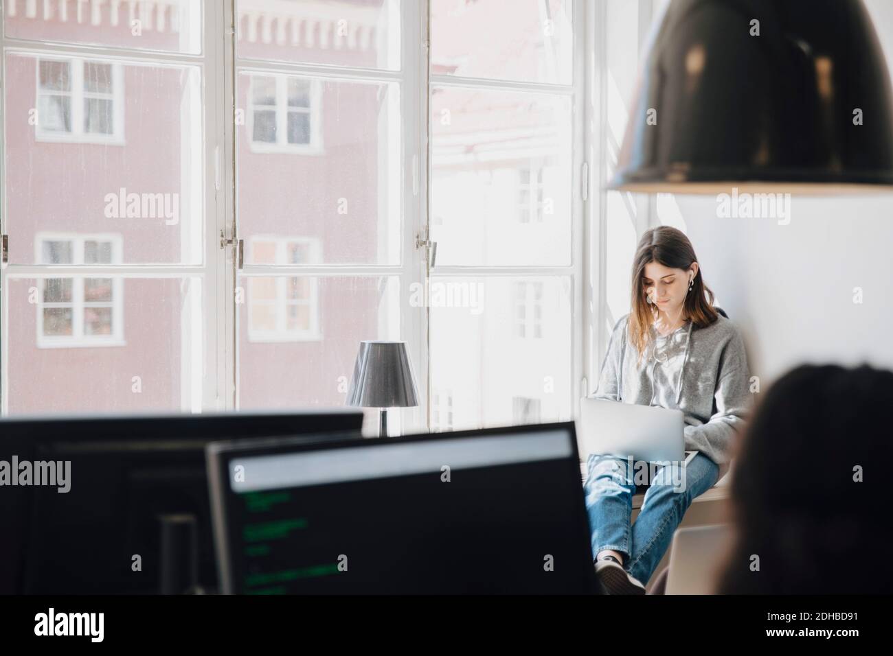 Focused young female programmer using laptop while sitting by window in ...