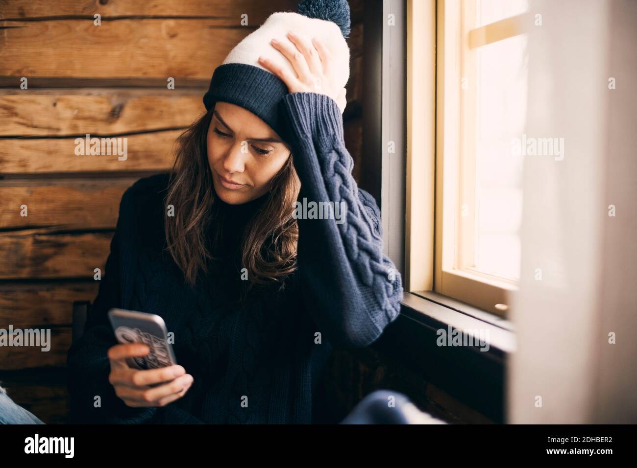 Mid adult woman using mobile phone while sitting by window in log cabin ...