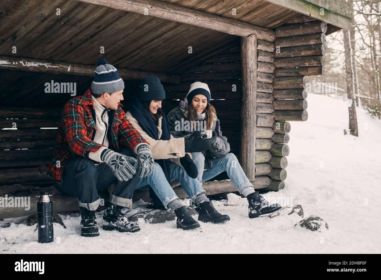 Friends sharing drink while sitting at log cabin during winter Stock ...