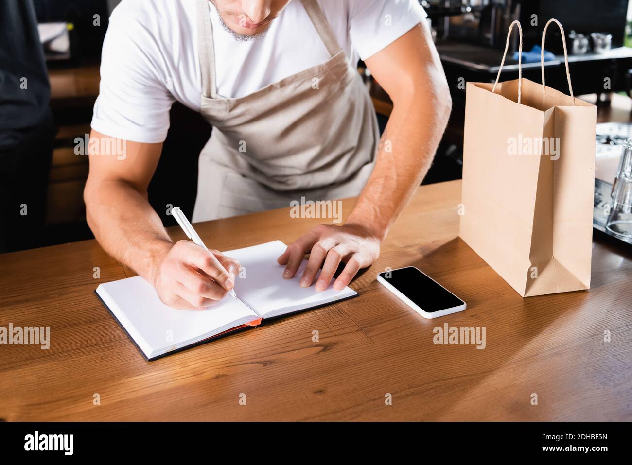 cropped view of barista writing in notebook near paper bag and ...
