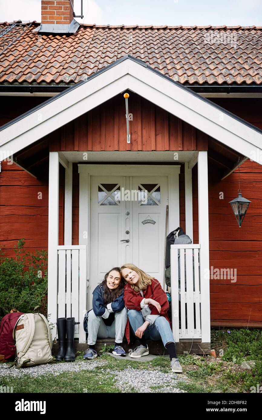 Portrait of smiling female friends with backpacks sitting outside ...