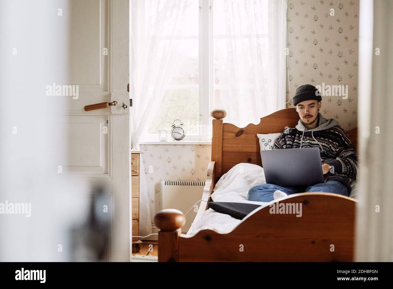Young man using laptop while sitting on bed seen through doorway at ...