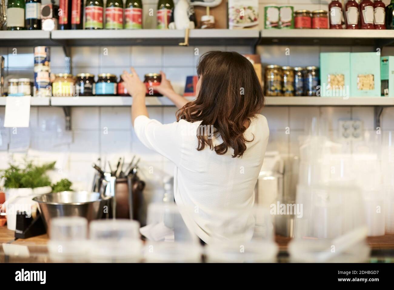 Rear view of female owner arranging food products on shelf in store ...