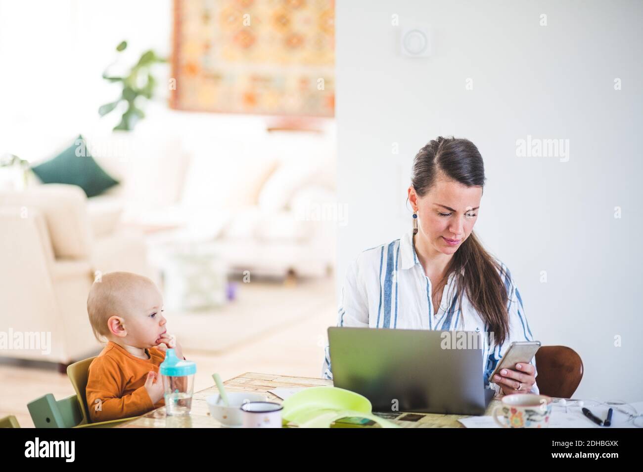 Baby girl looking at busy working mother while sitting at table in home ...