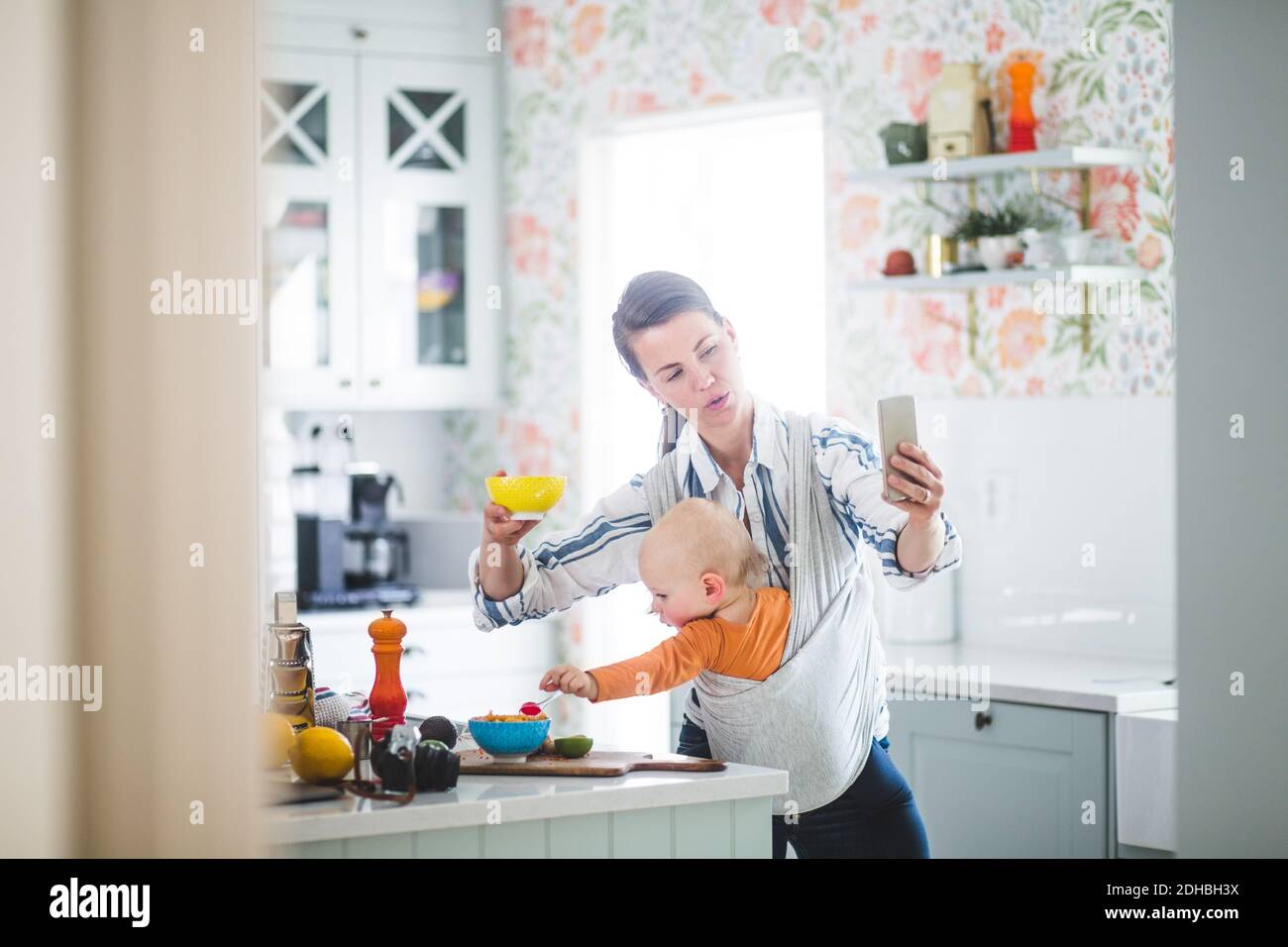 Multi-tasking mother food blogging while carrying daughter in kitchen ...