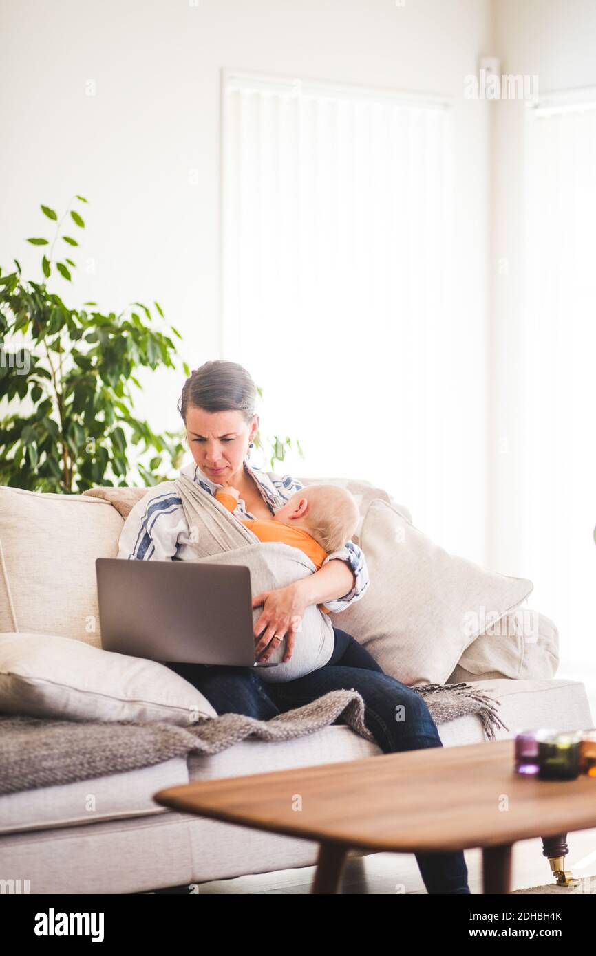 Multi-tasking mother using laptop while caring baby girl on sofa at ...