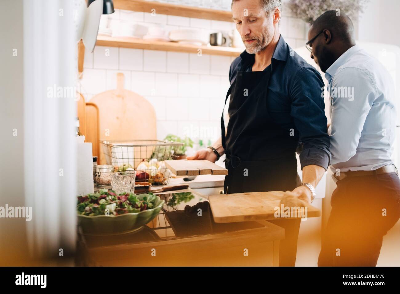 Mature man working at counter while standing with friend in kitchen ...