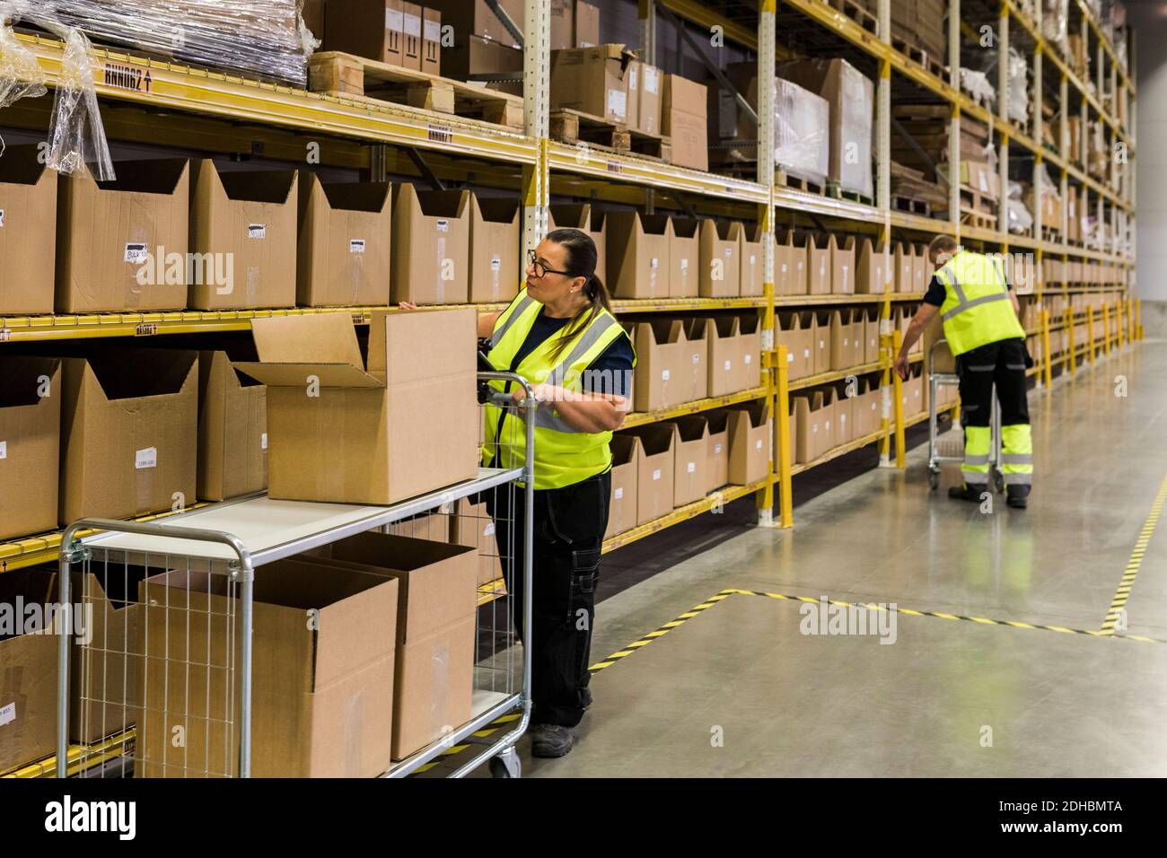 Full length of female worker pushing cart with cardboard boxes by rack ...