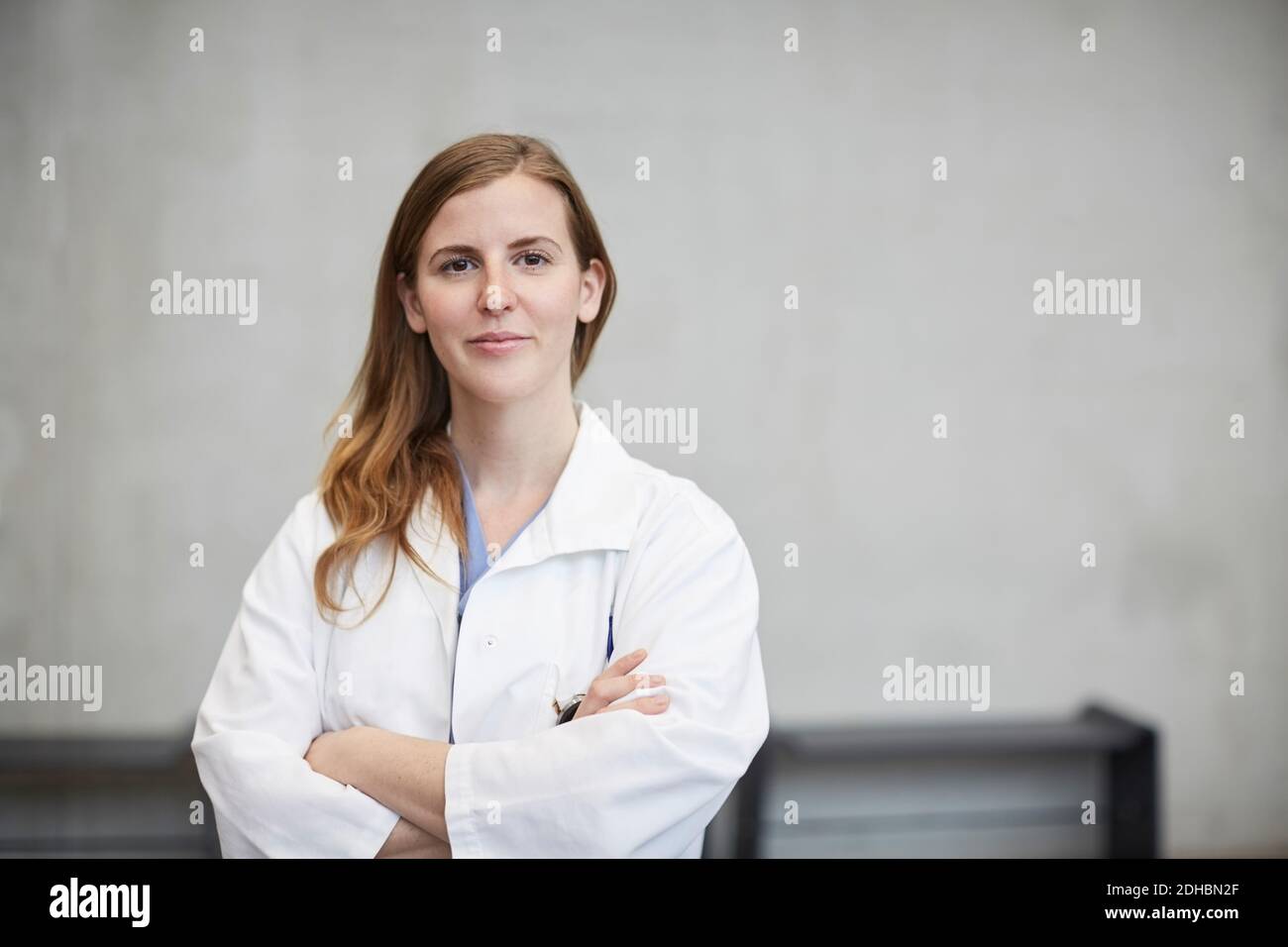 Portrait of confident mid adult female doctor standing with arms crossed against wall at ...