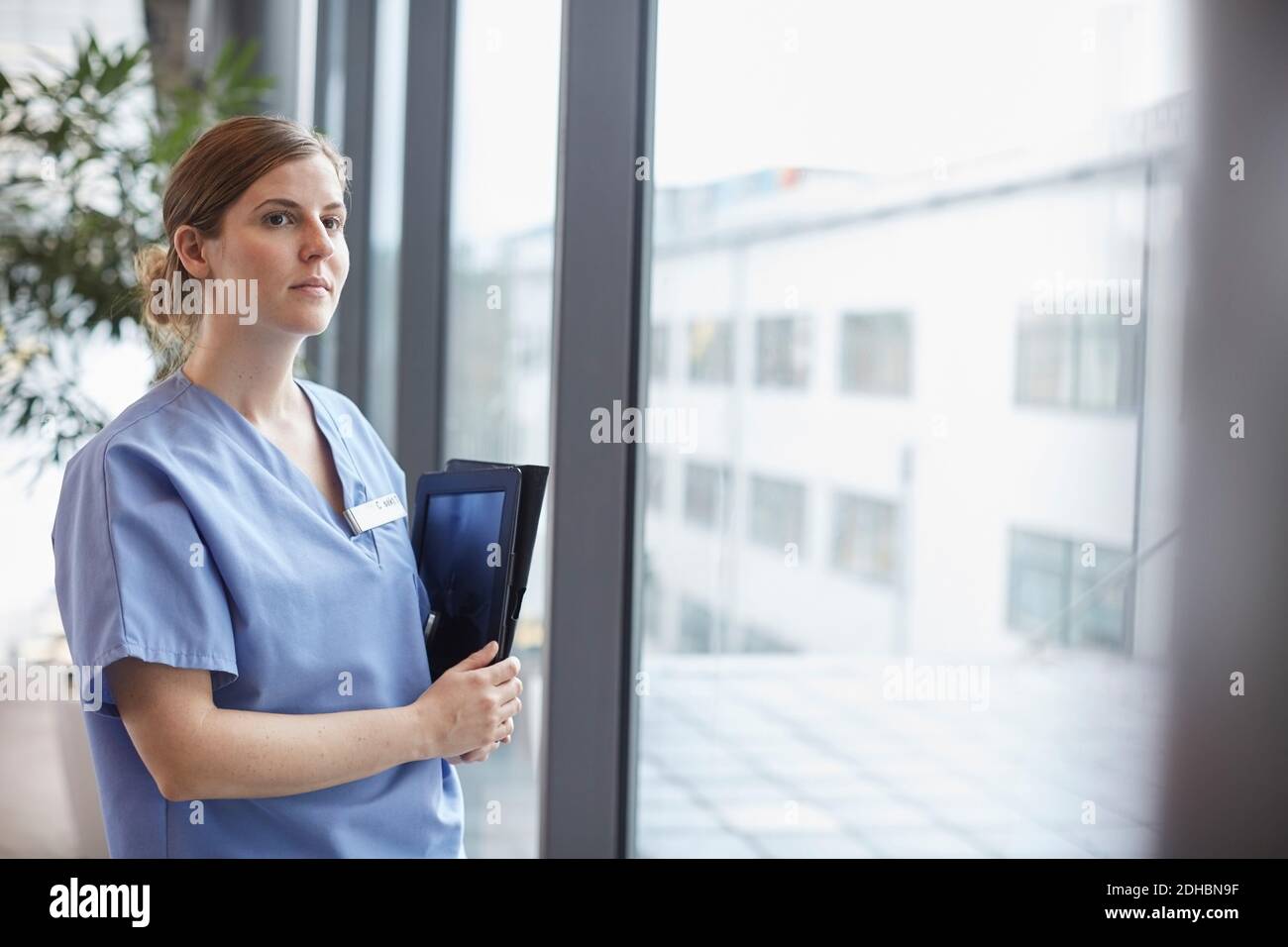 Thoughtful female nurse looking through window while standing in ...