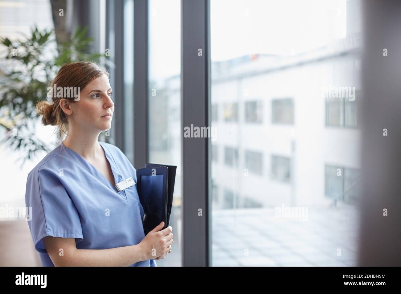 Thoughtful female nurse with digital tablet looking through window ...
