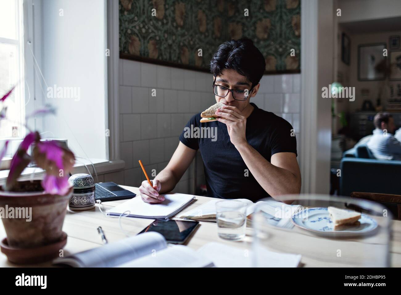 Young man eating sandwich while doing homework at table Stock Photo - Alamy
