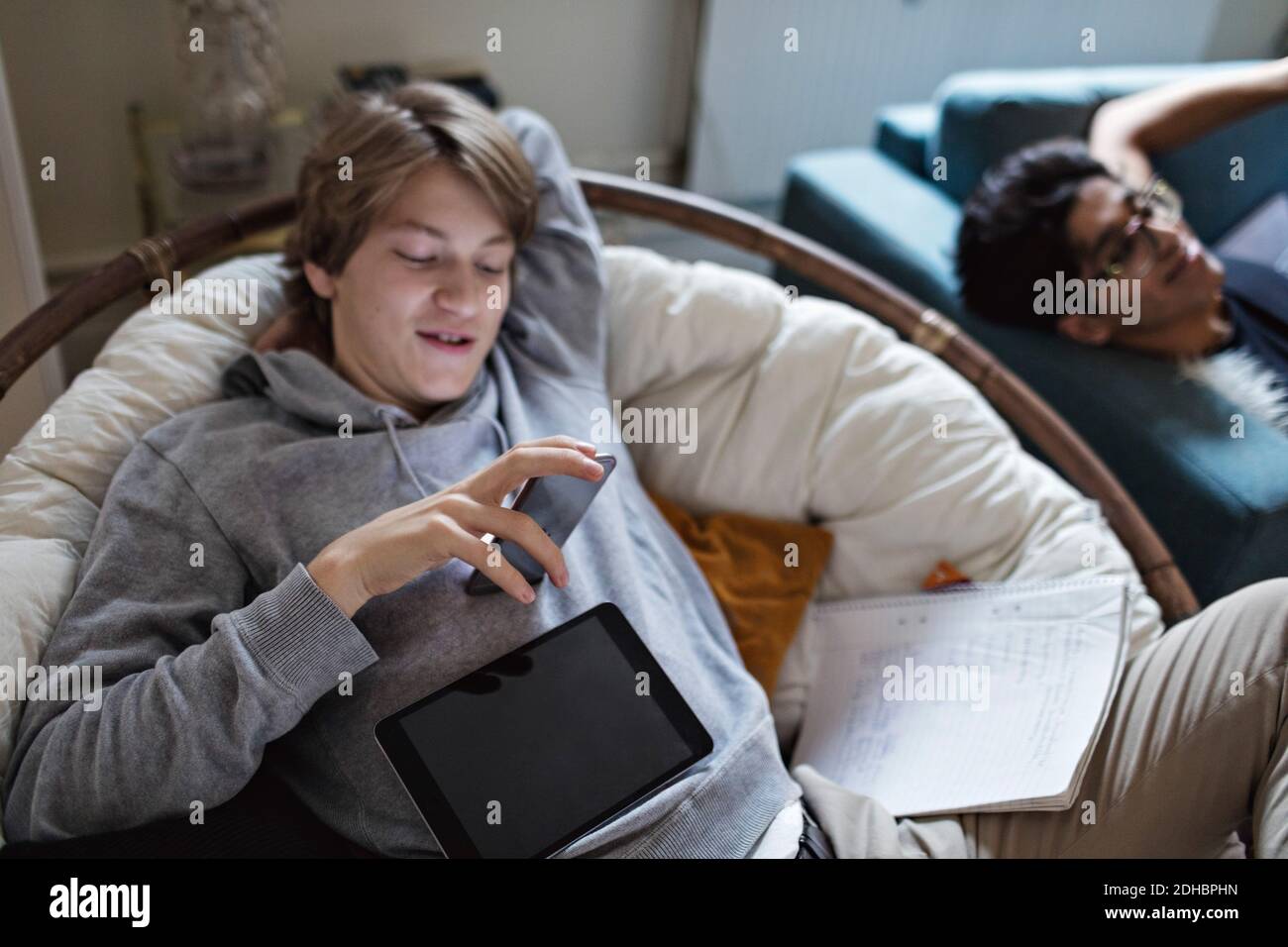 Teenage boy using mobile phone while studying with friend in living ...