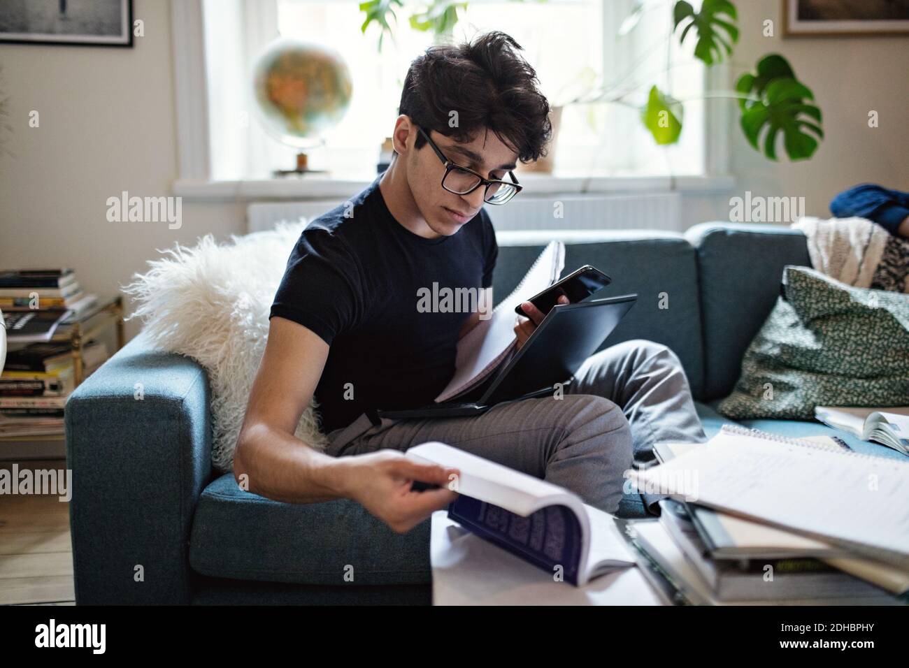 Serious young man doing homework while sitting on sofa with mobile ...