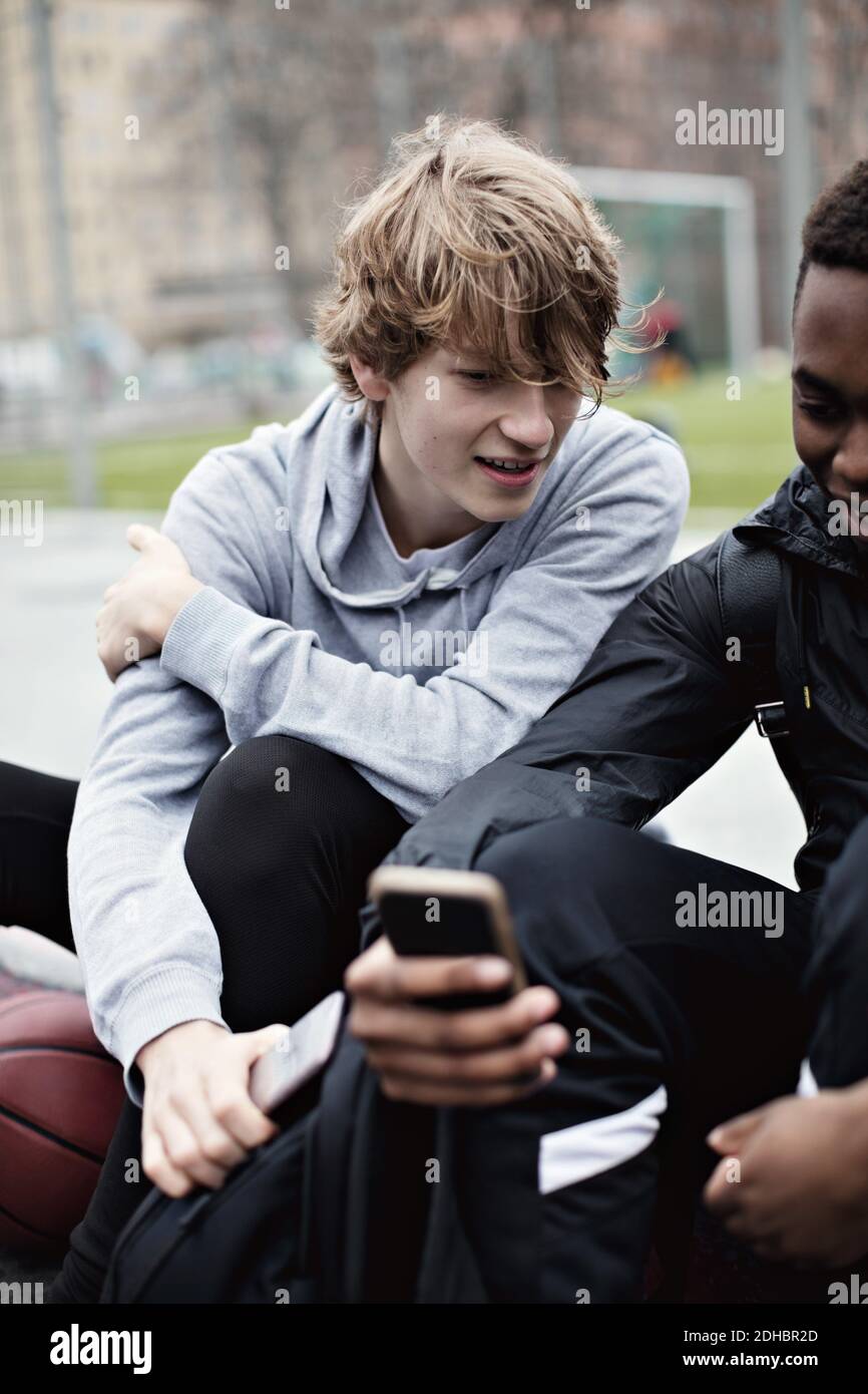 Teenage boy looking at friend's mobile phone while sitting on sidewalk ...
