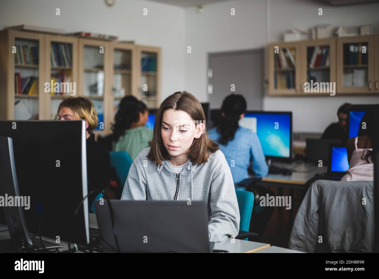 Confident female student using laptop at desk against teacher and ...