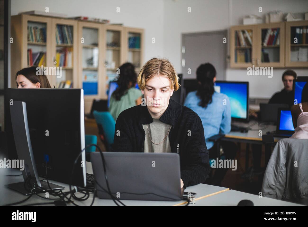 Young male high school student using laptop at desk against teacher and ...