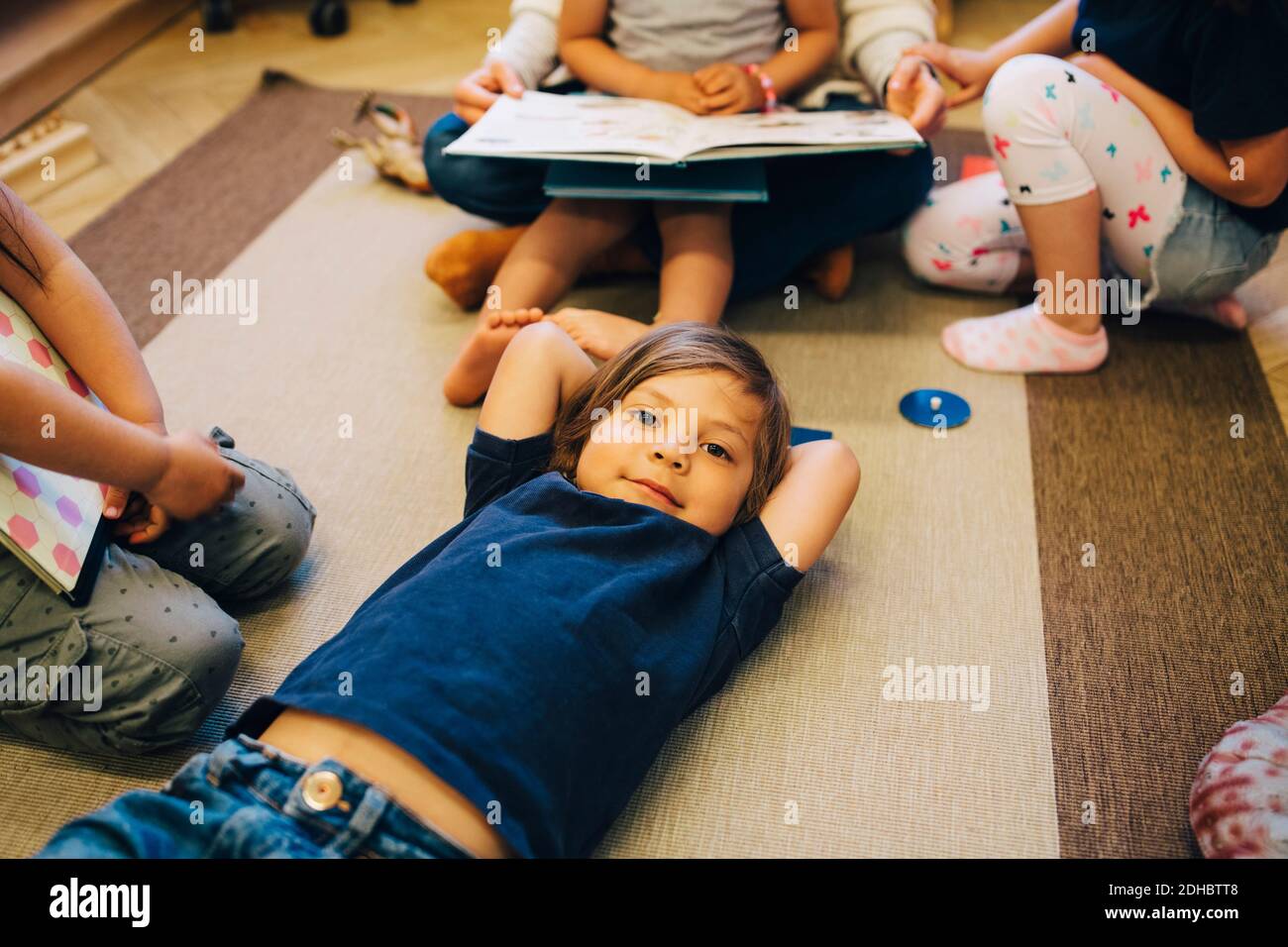 High angle portrait of boy lying on carpet amidst friends in classroom ...