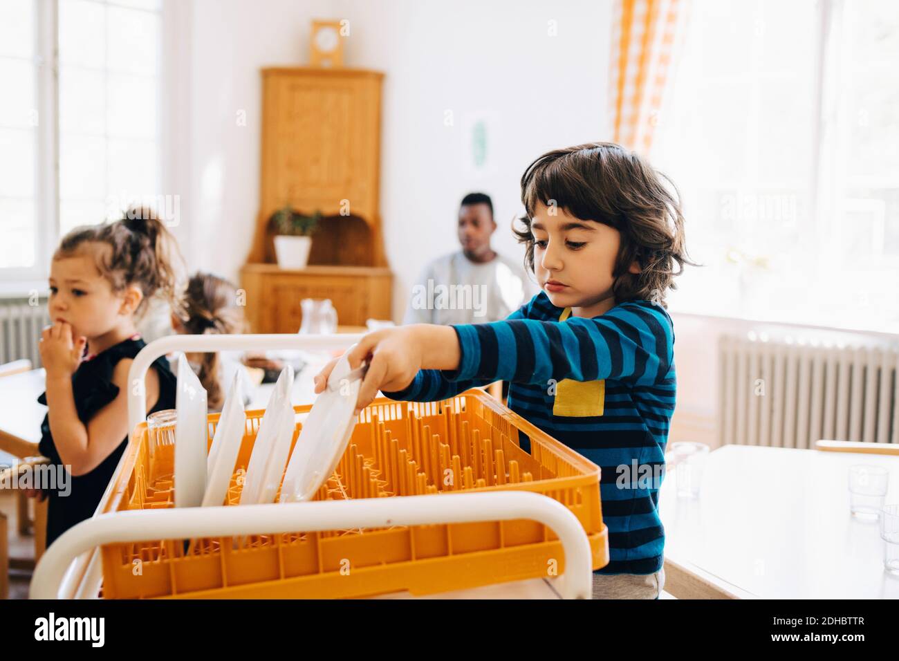 Boy placing plate in crate on cart by girl in classroom at kindergarten ...