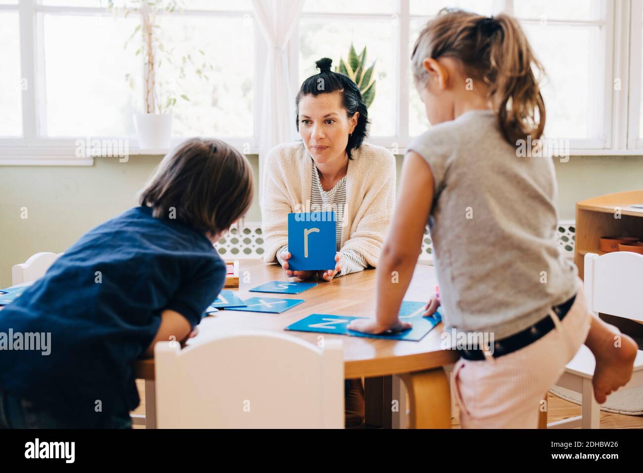 Confident female teacher holding letter R in front of students at table ...