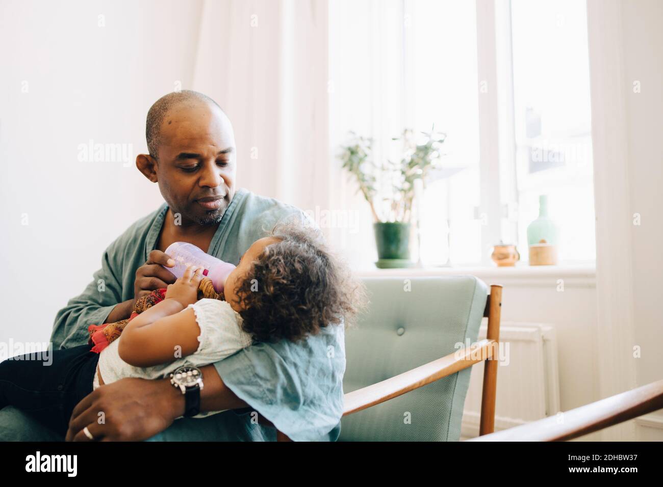 Caring father feeding milk to daughter while sitting in living room at ...