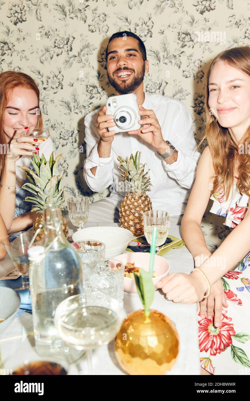 Smiling young man holding camera while sitting amidst female friends at ...
