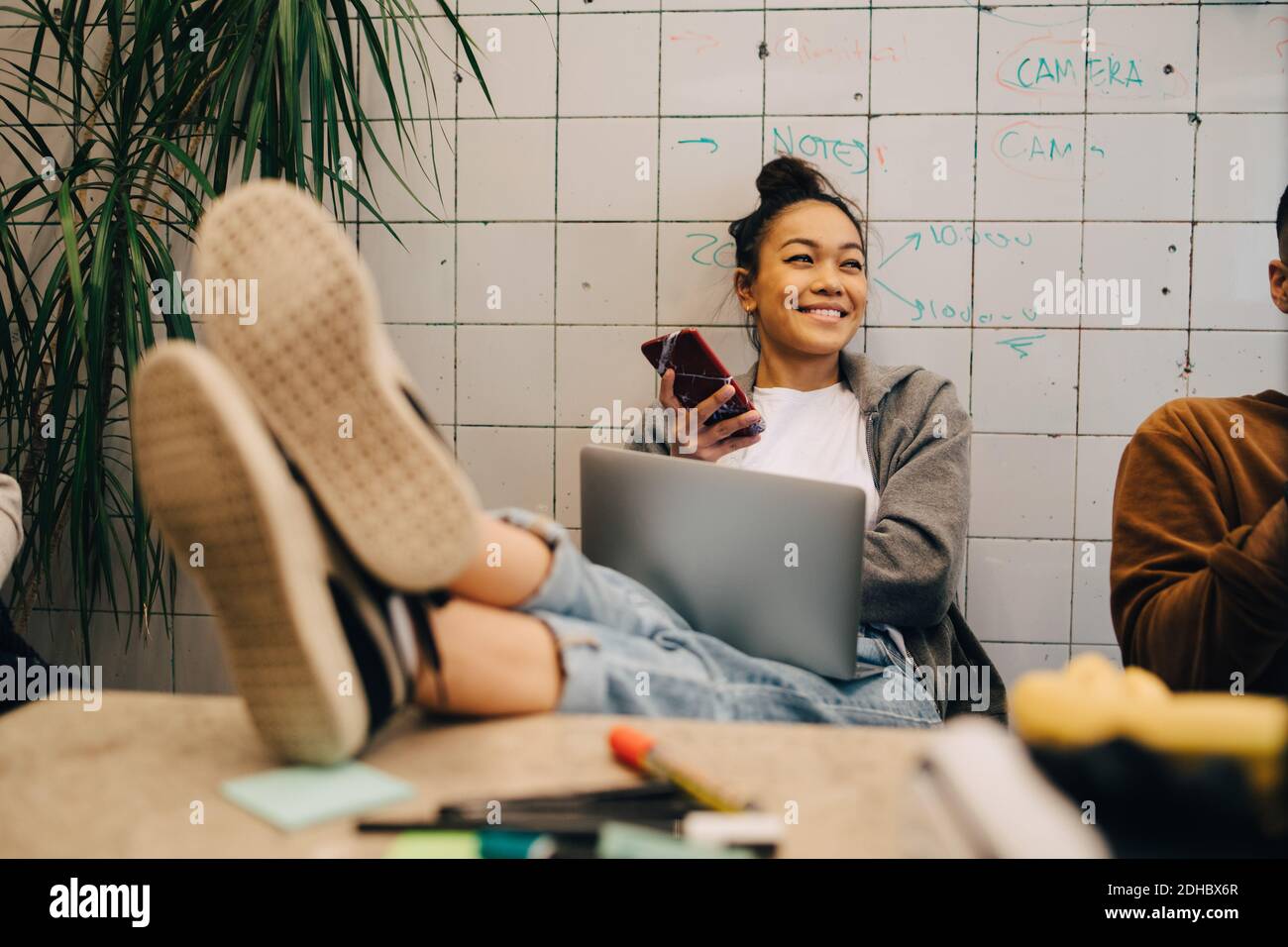 Smiling young businesswoman sitting with feet up on desk using wireless ...