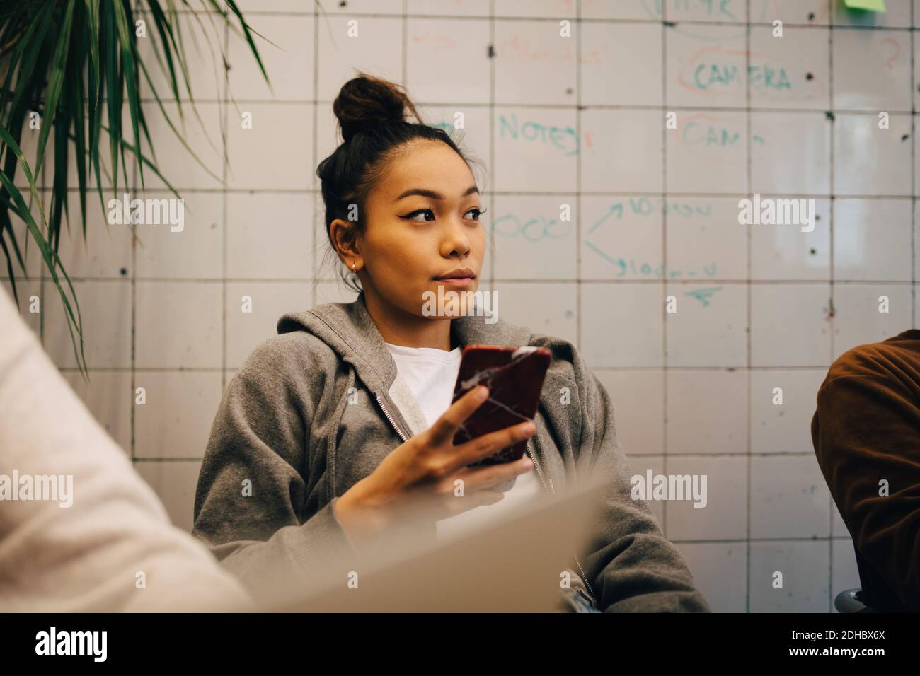 Young female programmer holding smart phone while sitting amidst ...