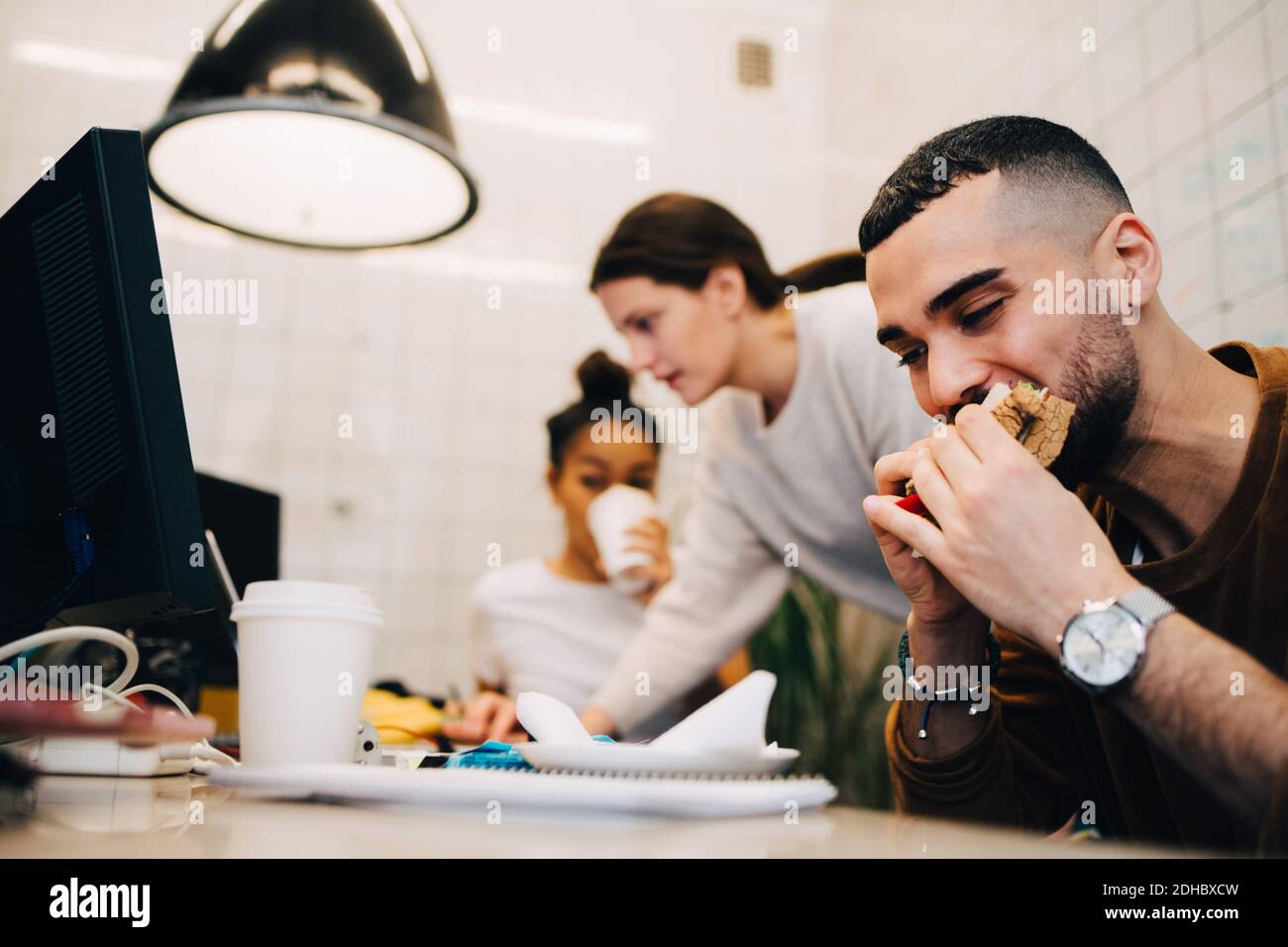Young male hacker eating sandwich while female colleagues working ...