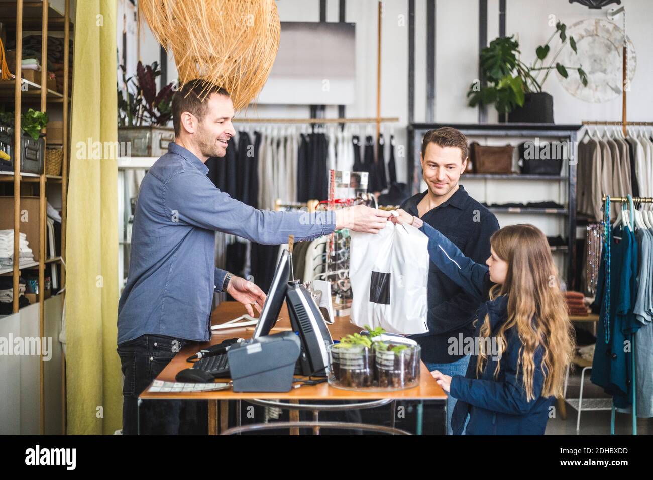 Smiling cashier giving shopping bag to girl by father in boutique Stock ...