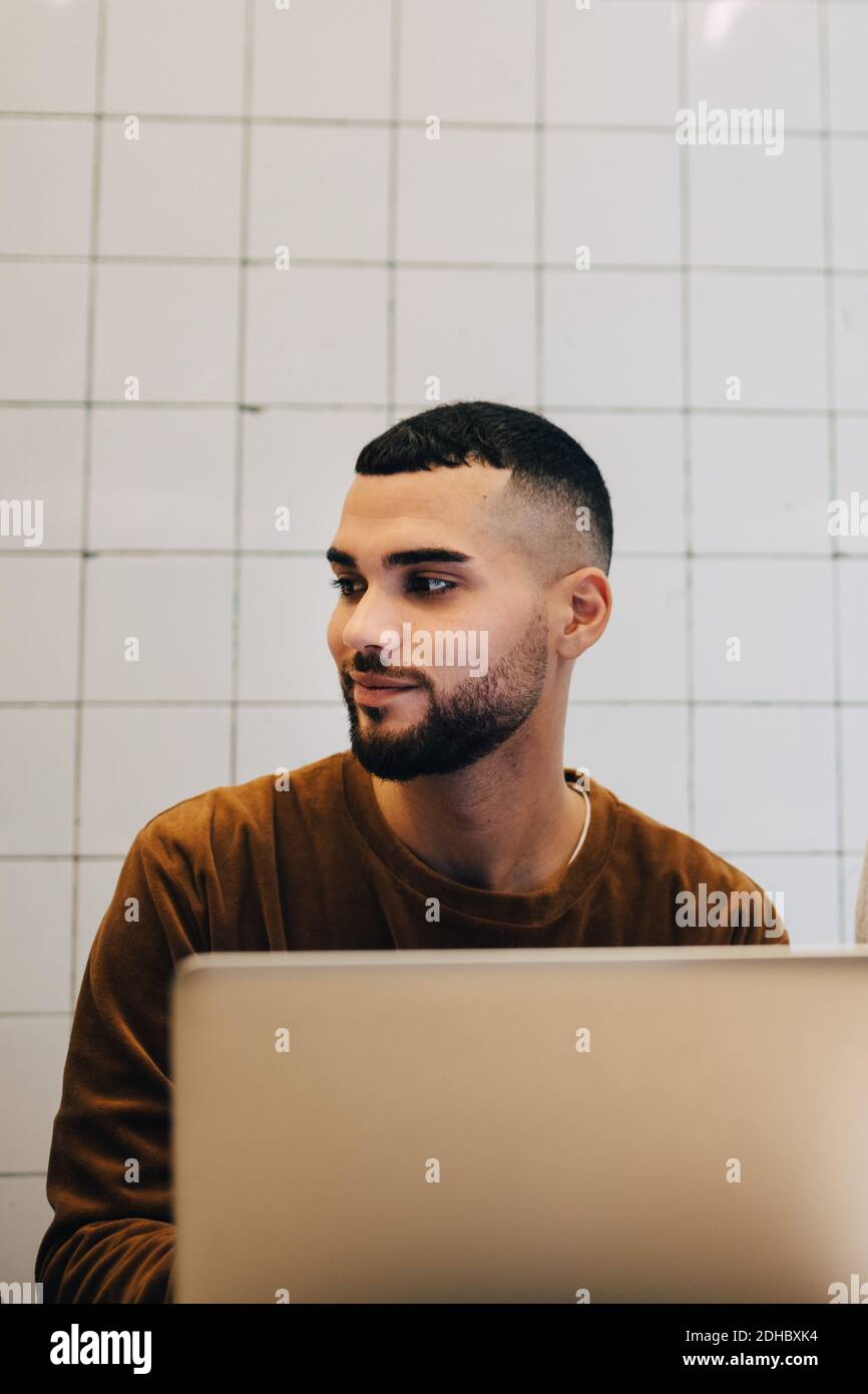Young hacker looking away while sitting with laptop against wall at ...