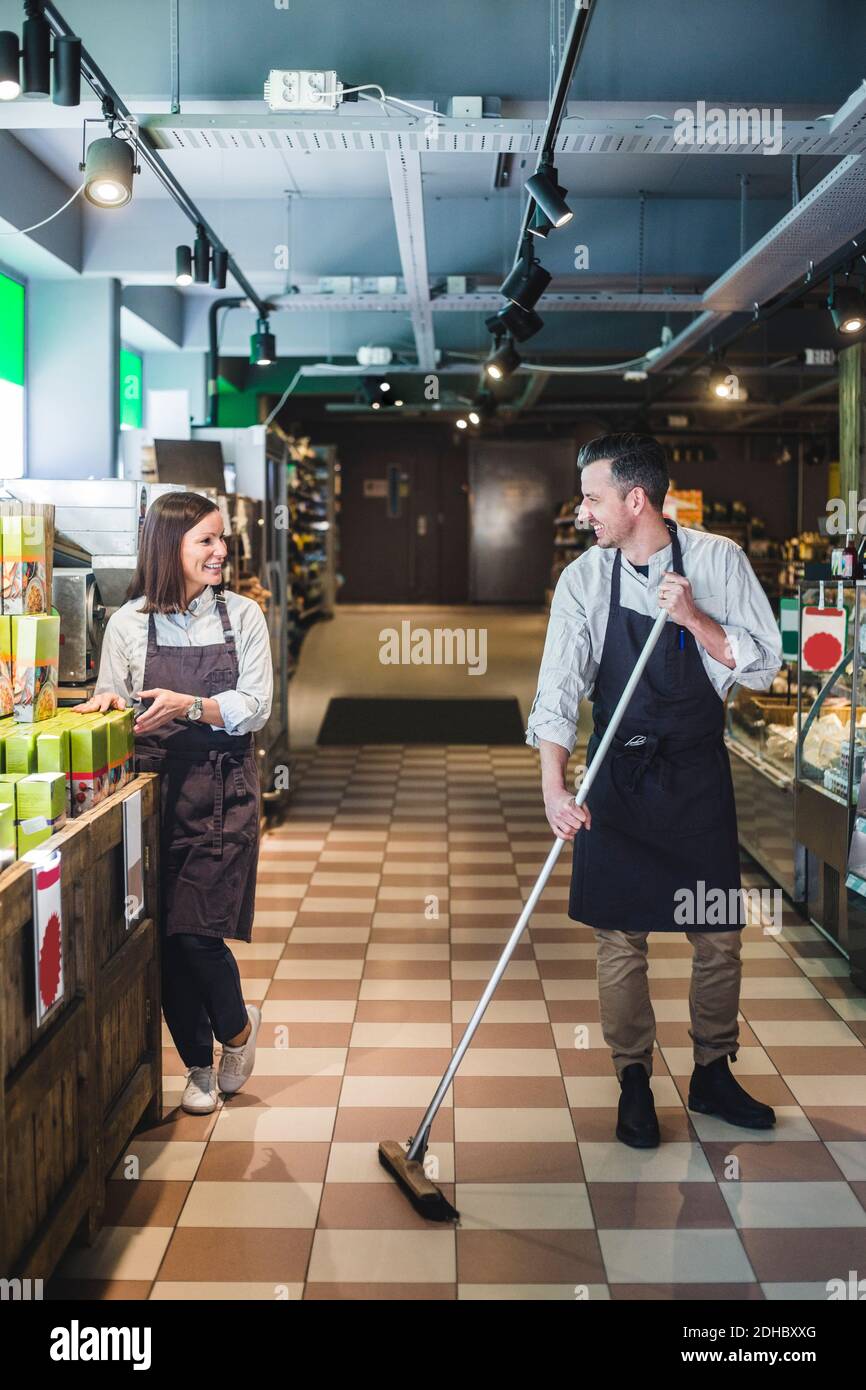 Smiling salesman sweeping floor while looking at colleague in ...