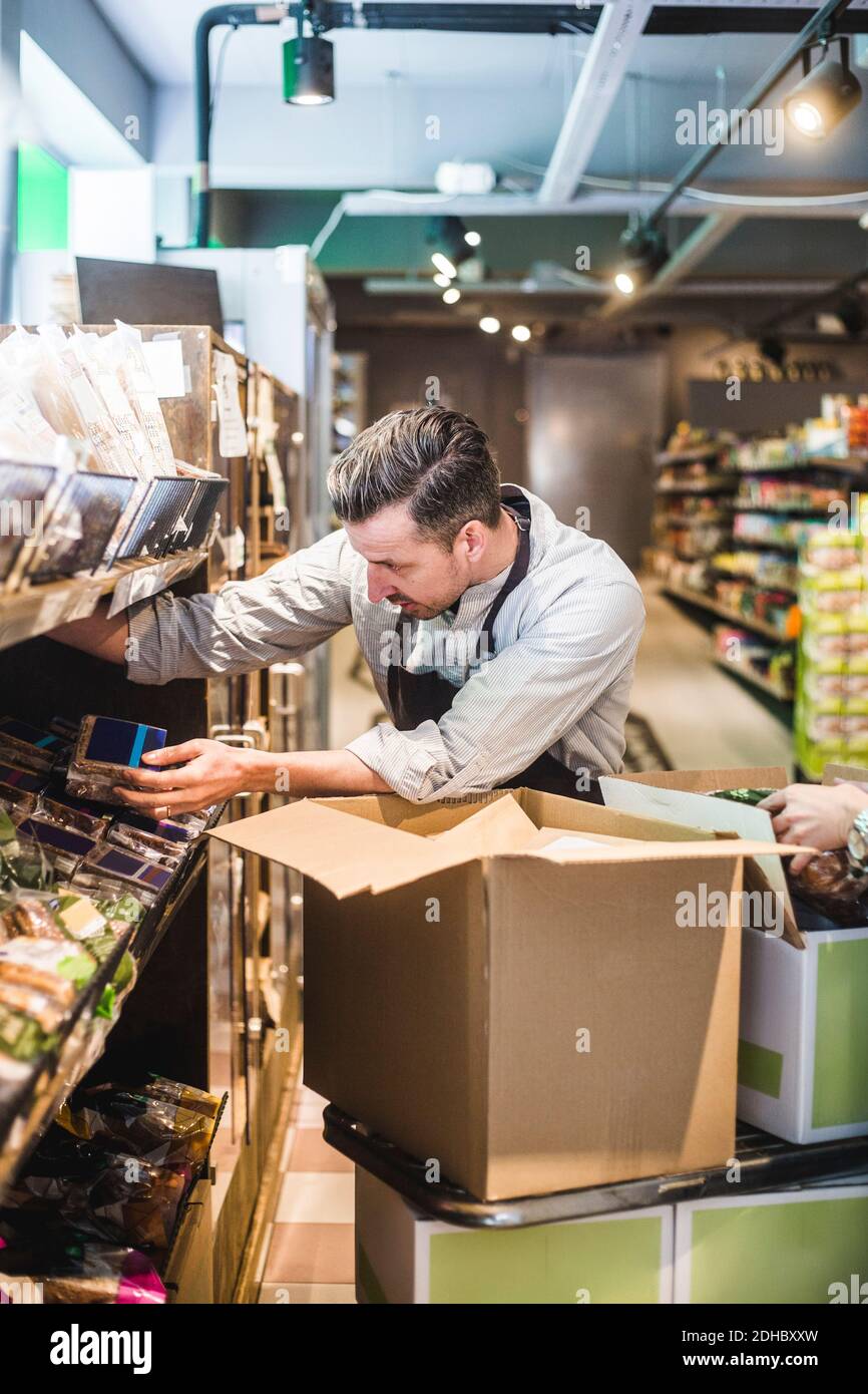 Salesman unloading cardboard box while arranging packets on shelf in ...