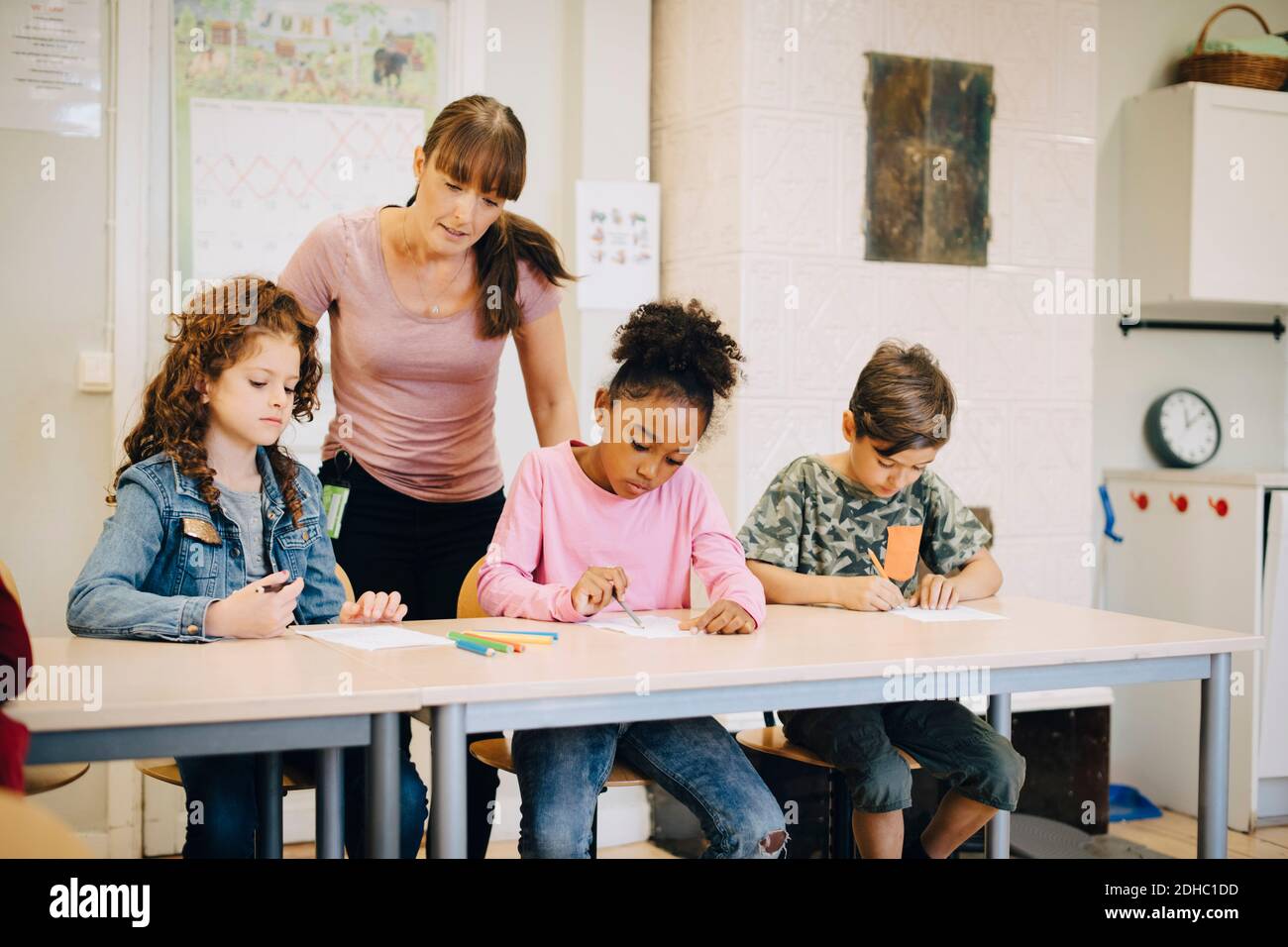 Teacher looking at students writing at desk in elementary classroom ...