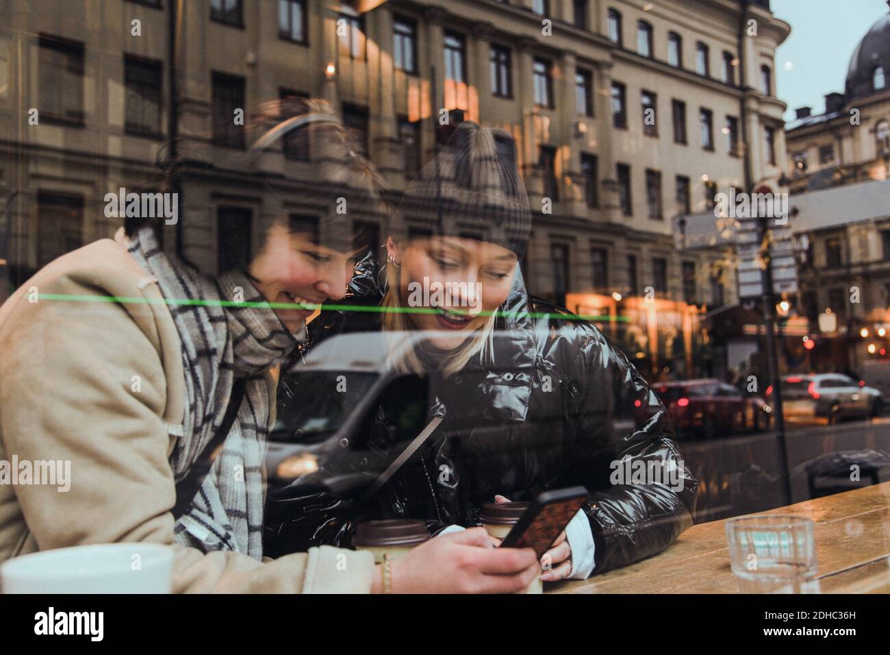 Smiling female friends using mobile phone while sitting in cafe seen ...