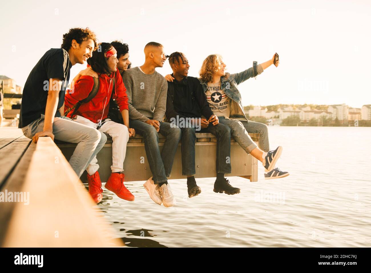 Happy male friends taking selfie while sitting side by side on pier by ...
