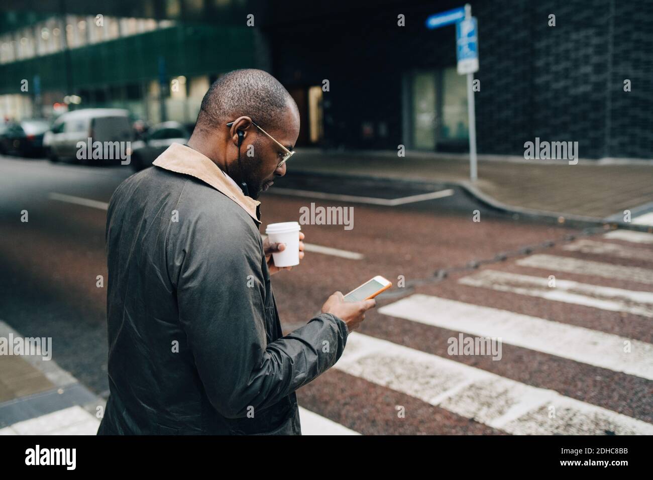 Side view of businessman with coffee using mobile phone while crossing ...