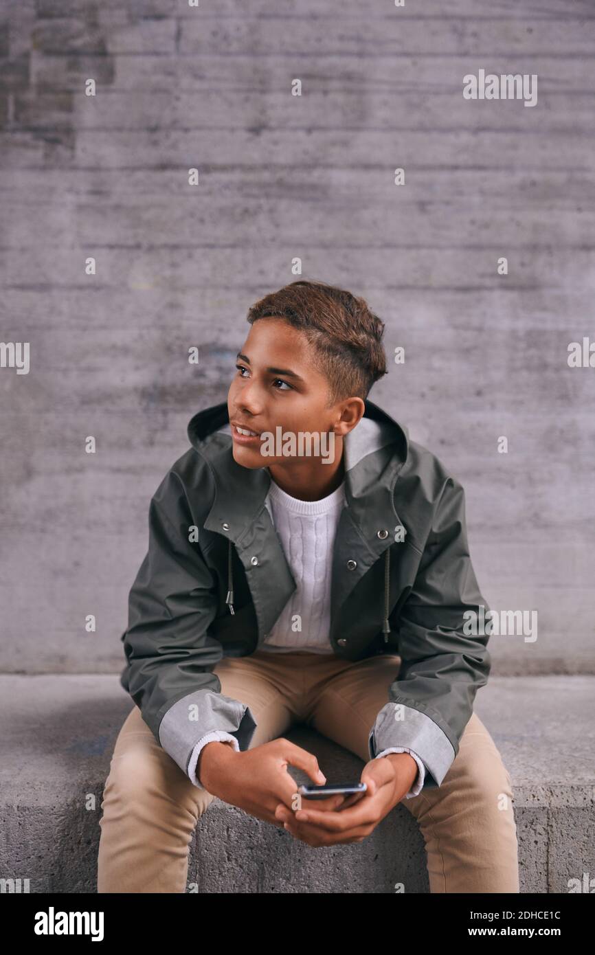 Smiling teenage boy with mobile phone looking away while sitting ...