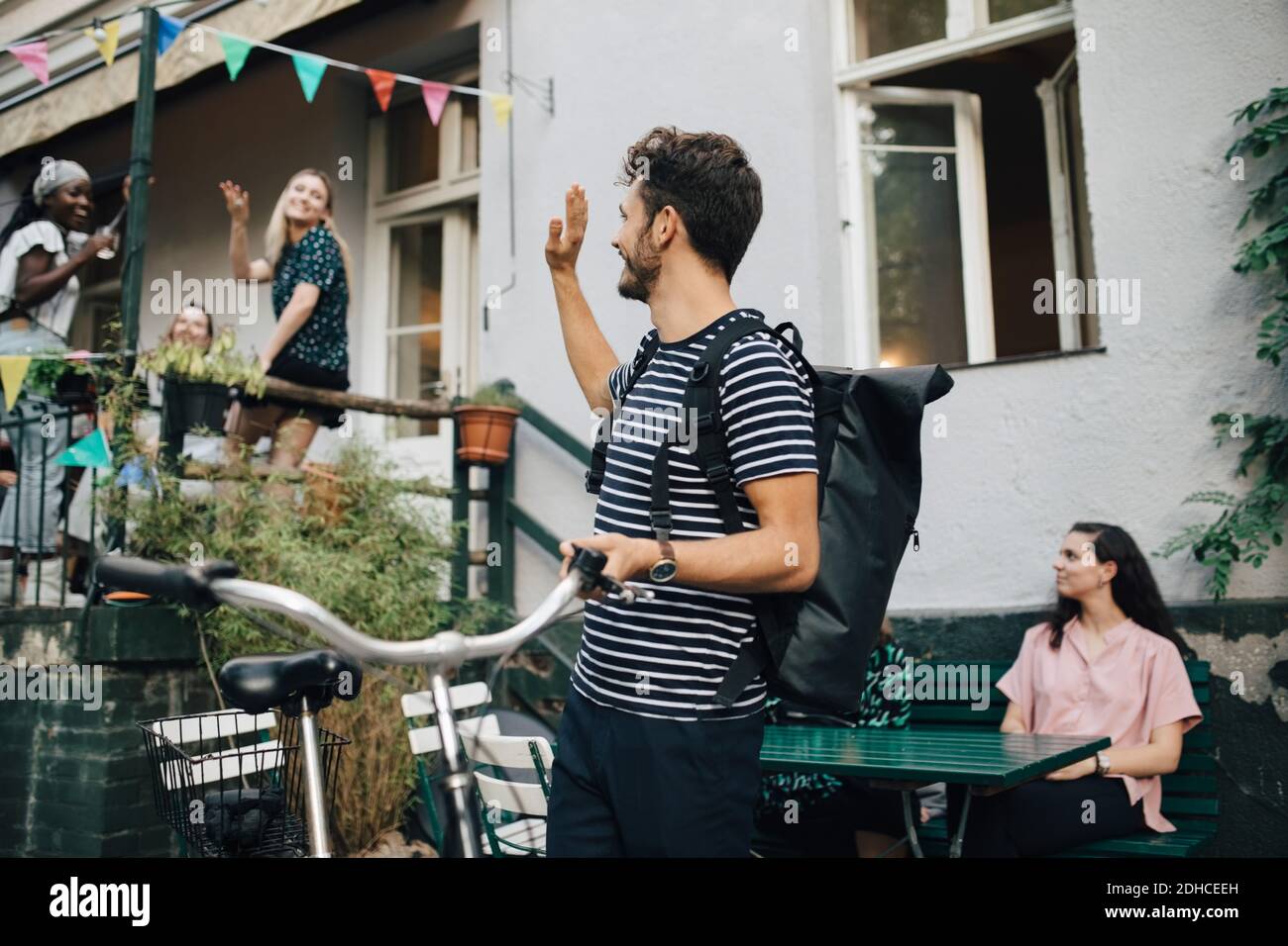 Smiling young man waving hand while standing with bicycle in backyard ...