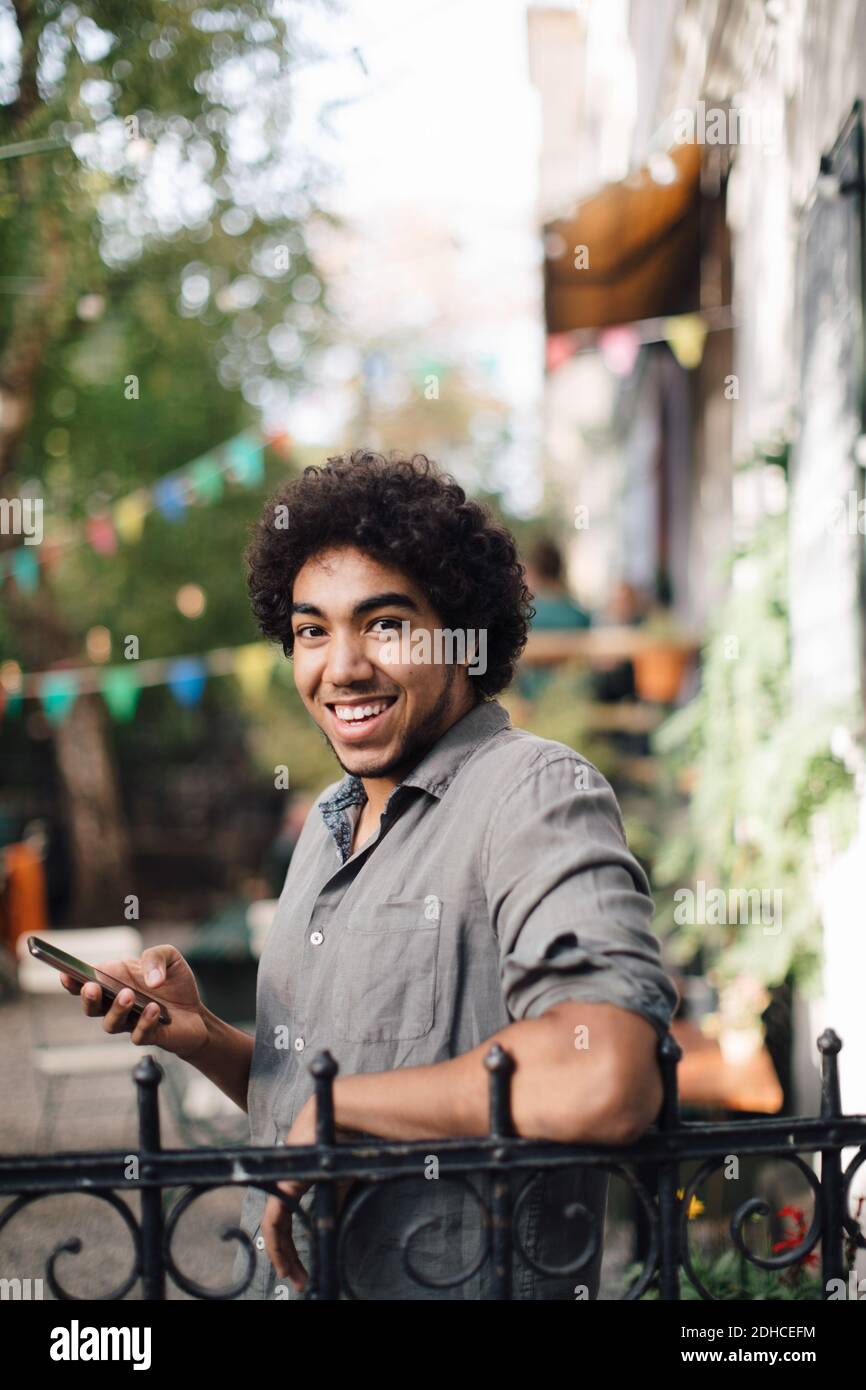 Portrait of smiling young man holding mobile phone while leaning on ...