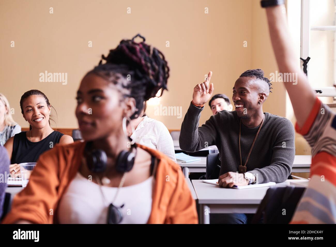 Happy young male student raising hand while sitting with classmates in ...