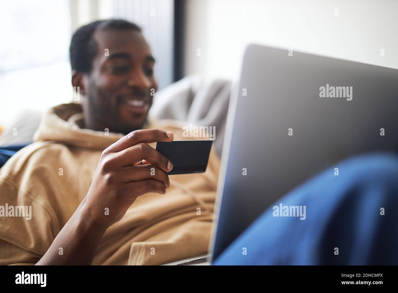 Side view of young mixed ethnicity man lying on sofa at home with laptop computer using credit ...