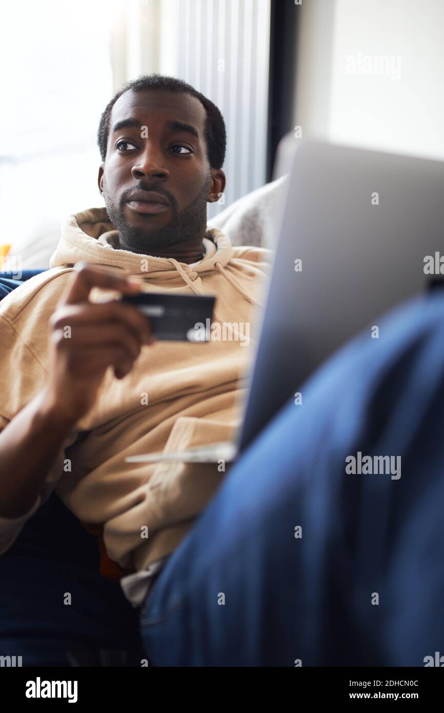Side view of young mixed ethnicity man lying on sofa at home with laptop computer using credit ...