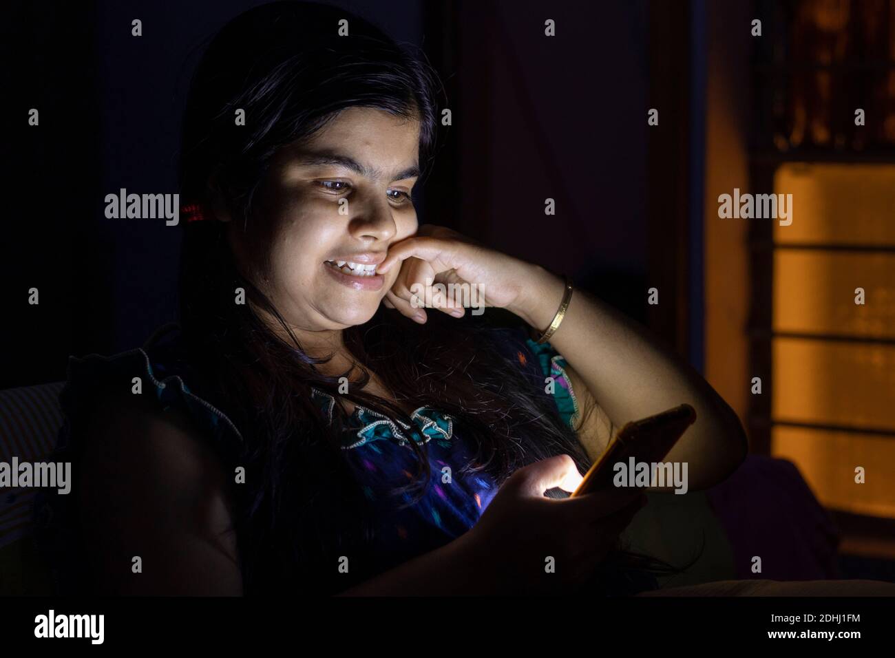 A Pretty Indian Woman Is Biting Nails While Looking At Smart Phone At Night In A Dark Room Stock