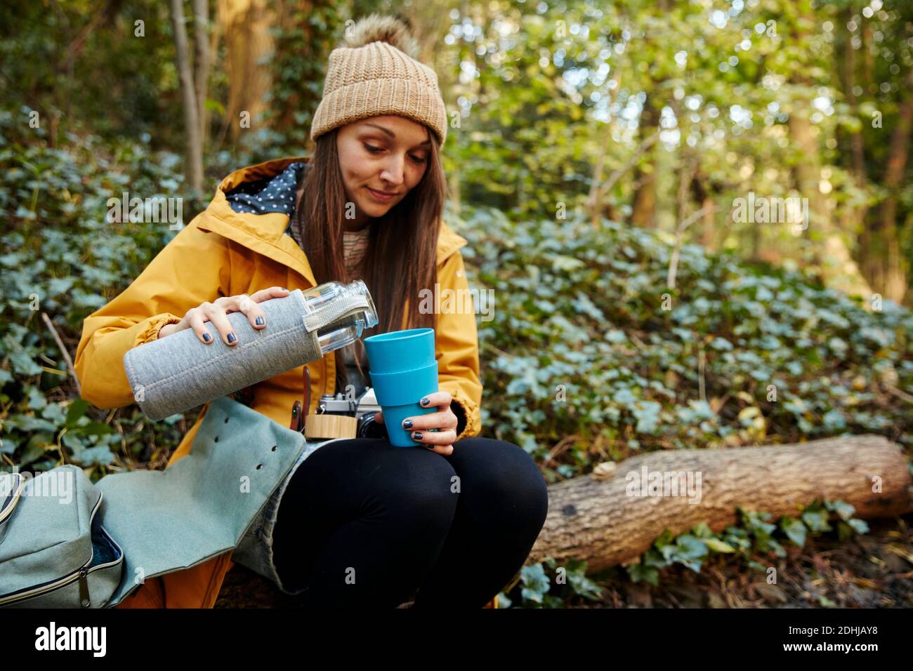 Woman sitting on tree trunk in woodland pouring drink from thermos ...