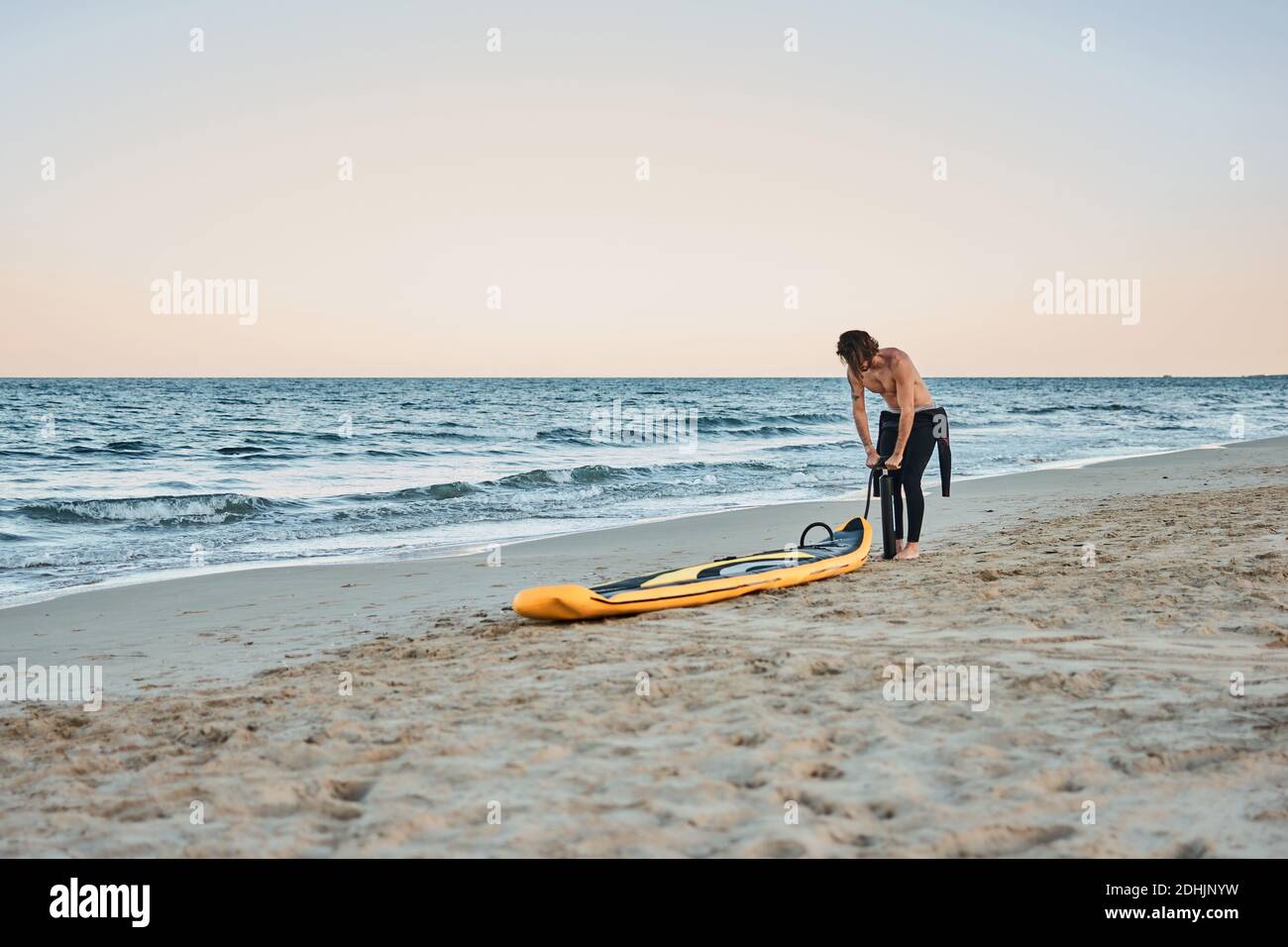 man in wetsuit inflating paddle board at the seashore on sandy beach ...
