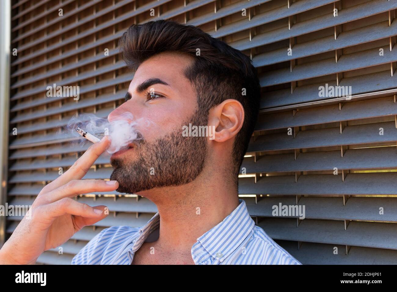Side view of thoughtful handsome male leaning on building and smoking ...