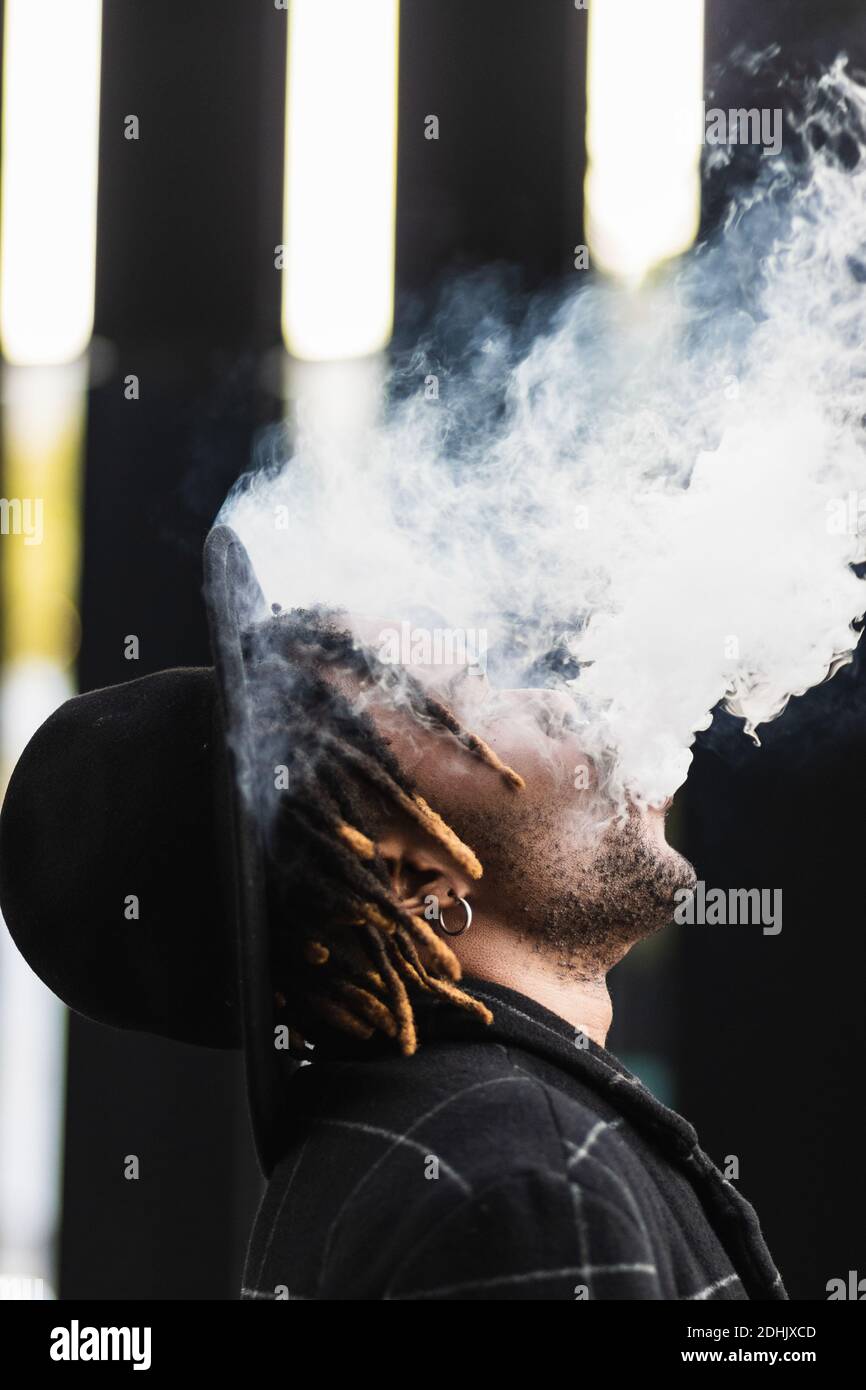 Side view of ethnic back guy with dreadlocks and hat in dense cloud of ...