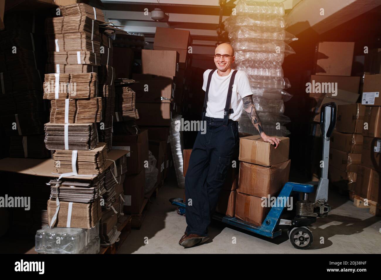 Smiling male warehouse worker leaning on stack of carton boxes on ...
