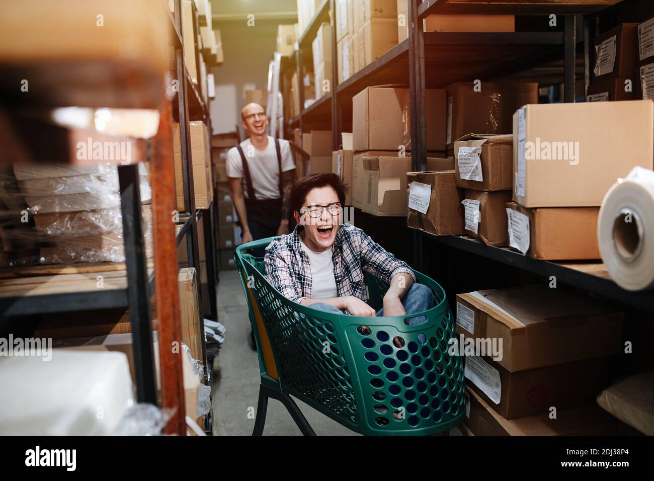 Warehouse workers having fun, man pushing cart with woman inside and ...